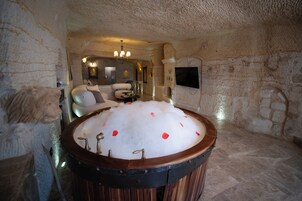 Quarto com paredes de pedra, uma banheira grande cheia de água e pétalas de rosa, um sofá e uma televisão fixada na parede.
