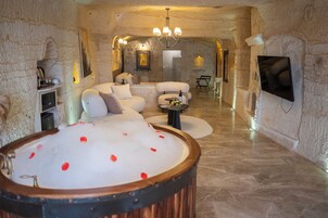 Um quarto de spa com interior em pedra, uma banheira grande com pétalas de rosa, um sofá e uma televisão.