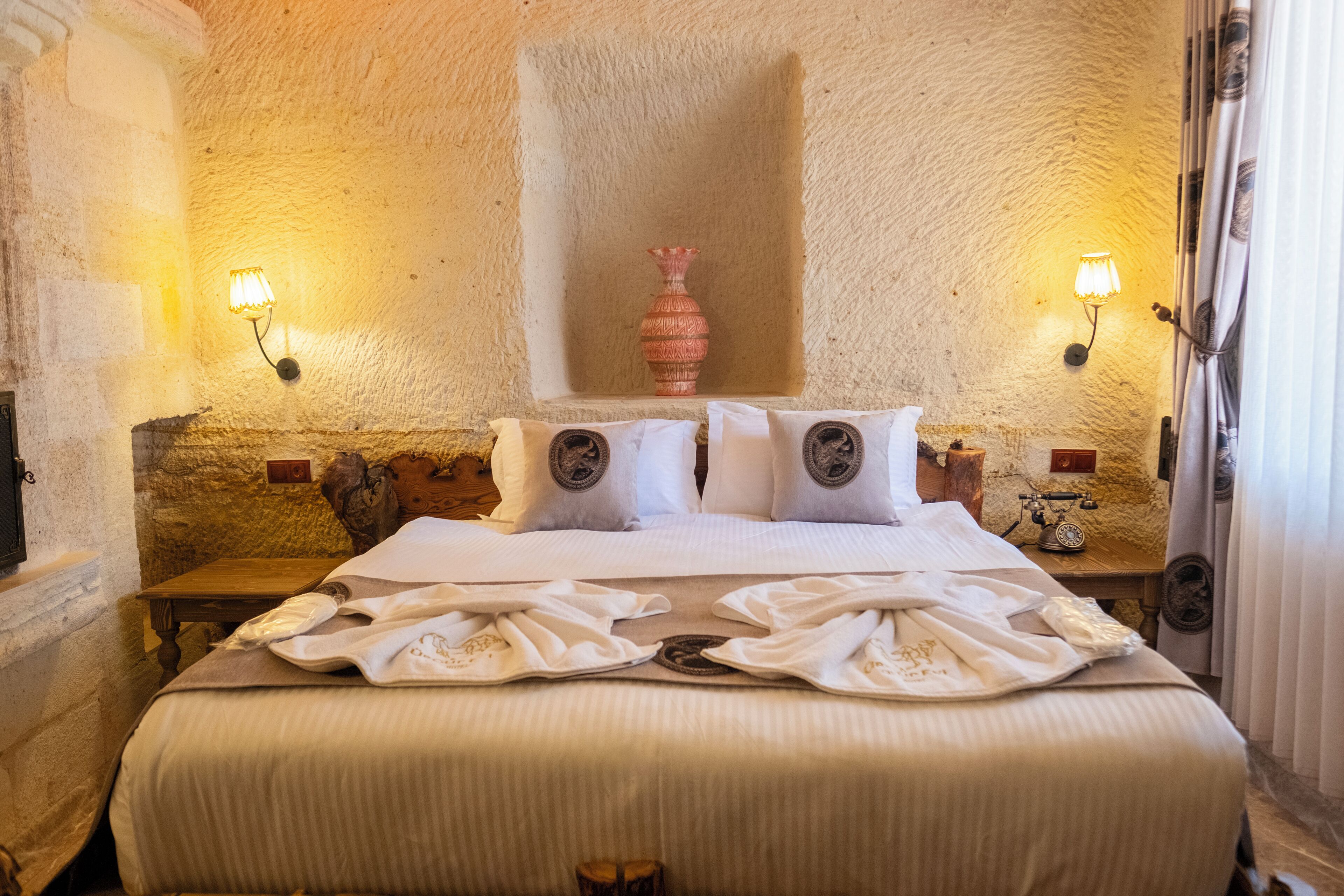 Quarto de hotel com uma cama grande, duas mesas de cabeceira, um painel de cabeceira de madeira, um vaso decorativo e luminárias fixadas na parede.