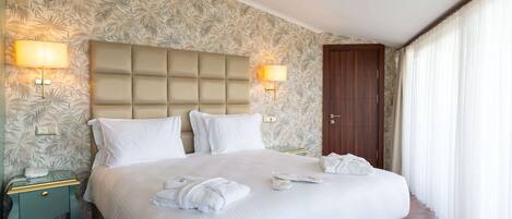 Quarto de hotel com uma cama grande, duas luminárias de cabeceira, um guarda-roupa de madeira e uma janela com cortinas translúcidas.