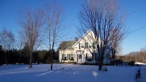 Uma casa de dois andares com quintal coberto de neve e árvores sem folhas.