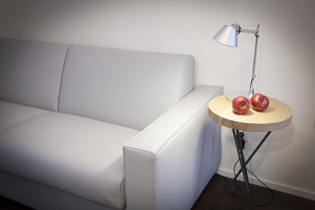 Um sofá branco com uma mesa lateral de madeira redonda, onde estão duas maçãs e um abajur.