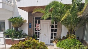 Entrada de prédio com porta branca, placa de 'PERIGO NÃO ENTRE' e diversos vasos de plantas.