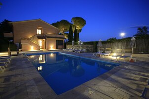 Piscina com espreguiçadeiras e guarda-sóis, escadaria que leva a um edifício e um sebedo ao fundo.