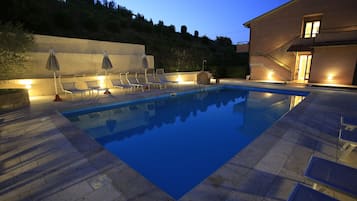 Piscina com espreguiçadeiras e guarda-sóis, cercada por um deck de pedra e um edifício com varanda.