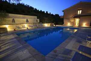 Piscina com espreguiçadeiras e guarda-sóis, cercada por um deck de pedra e um edifício com varanda.