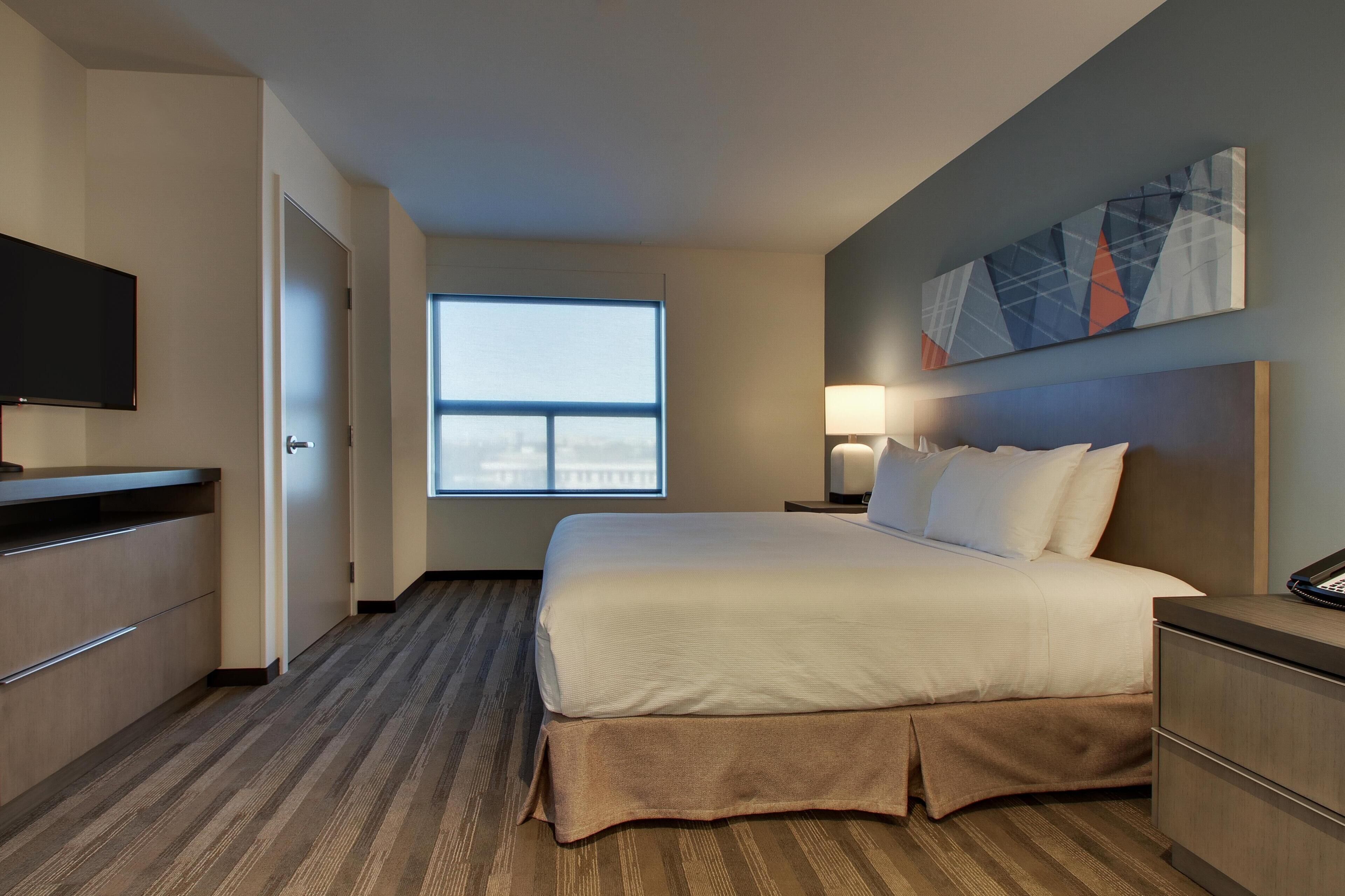 Quarto de hotel com cama, mesa de cabeceira, luminária, televisão e janela.