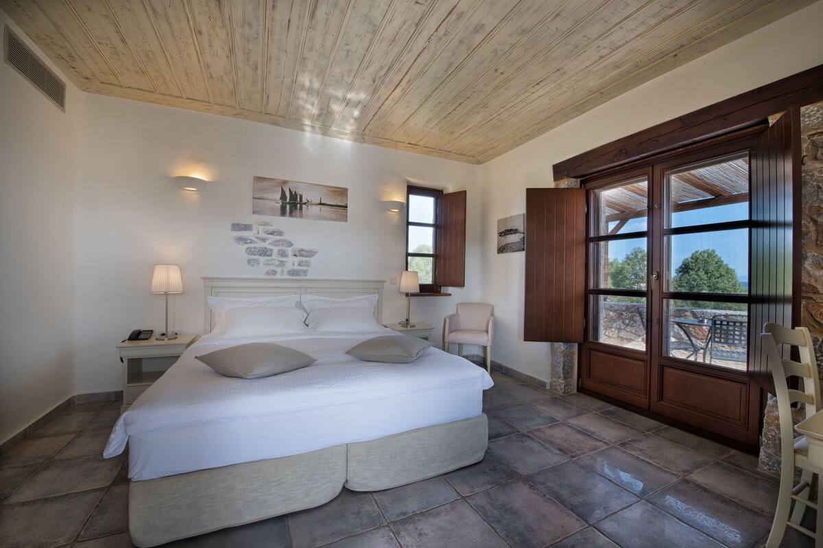 Quarto com uma cama grande, duas mesas de cabeceira, uma cadeira e janela com vista.