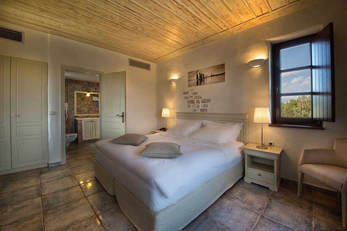 Quarto com cama grande, mesas de cabeceira, uma janela e um banheiro visível por uma porta aberta.