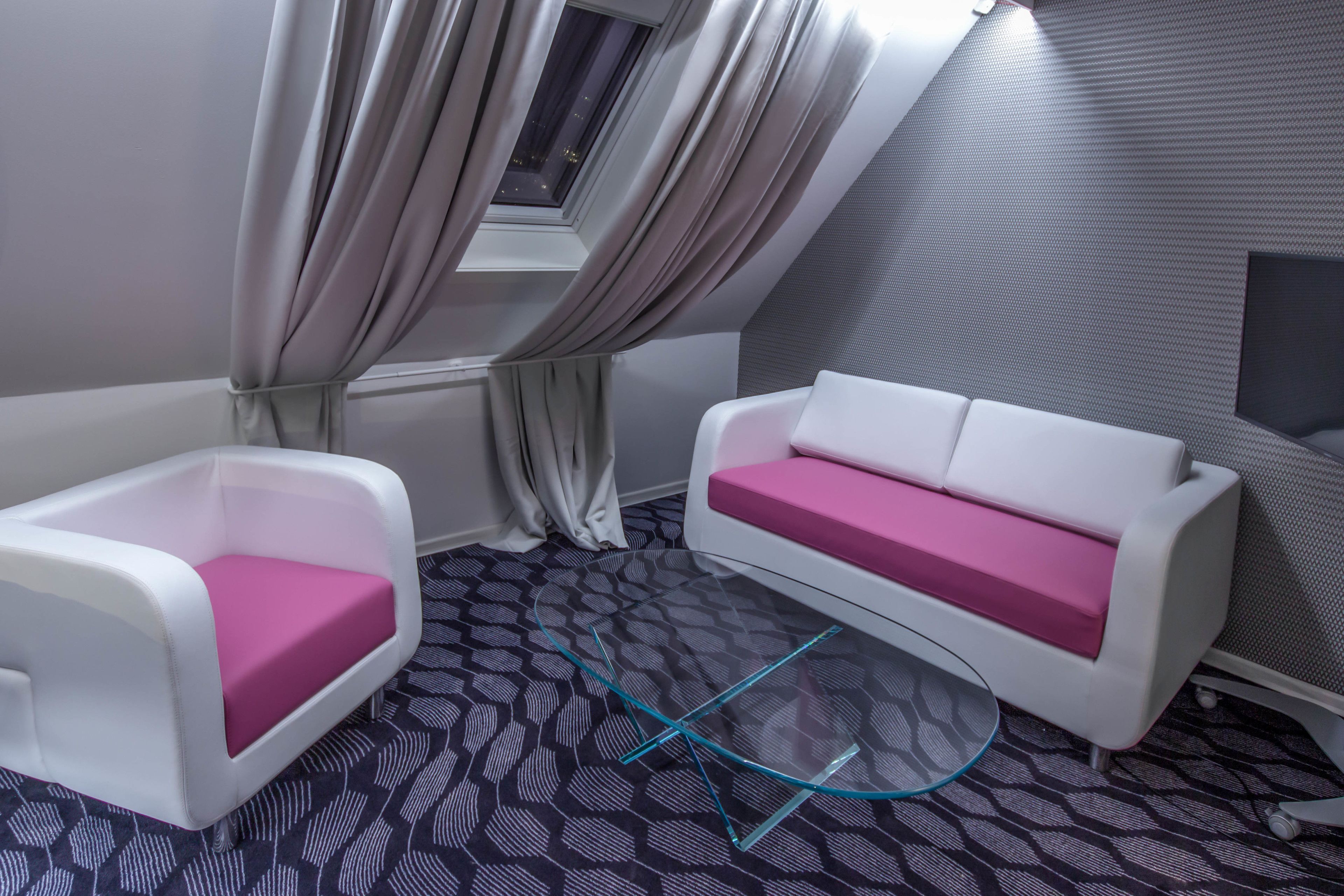 Quarto com sofá branco, almofadas cor-de-rosa, poltrona branca, mesa de centro de vidro, tapete estampado e janela com cortinas.