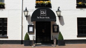 Entrada do The Rugby Hotel, com um arco preto, duas luminárias estilo lanterna também pretas e caixas de flores nas janelas.