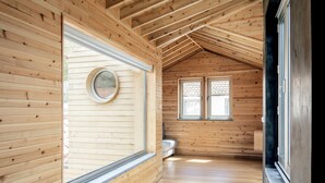 Interior de uma cabana de madeira com teto inclinado, janelas amplas e soalho em madeira.