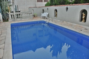Piscina com deck de pedra, um banco branco e estátuas.