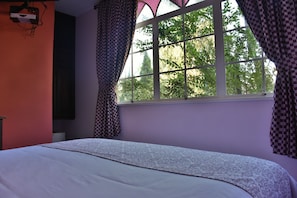 Quarto com cama, janela com cortinas xadrez e vista para árvores lá fora.