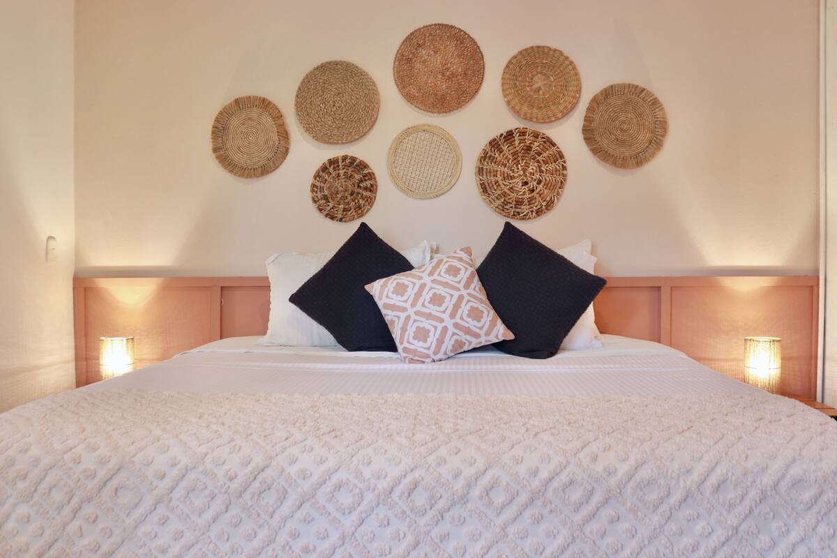 Uma cama bem arrumada com um edredom estampado, duas almofadas pretas e uma com padrão geométrico. Nove peças de tapeçaria adornam a parede acima da cama.