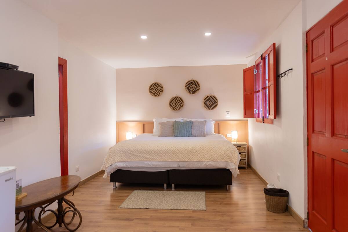 Quarto com cama, mesas de cabeceira, televisão e porta vermelha.