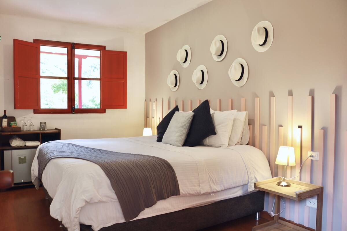 Quarto bem organizado com uma cama grande, mesa de cabeceira com luminária, guarda-roupa e janela com persianas vermelhas.