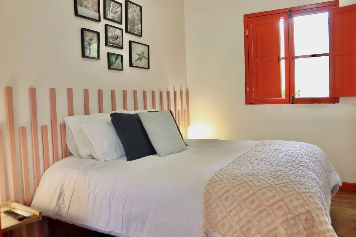 Cama bem arrumada com edredom branco e almofadas, mesa de cabeceira com controle remoto e janela com persianas vermelhas.