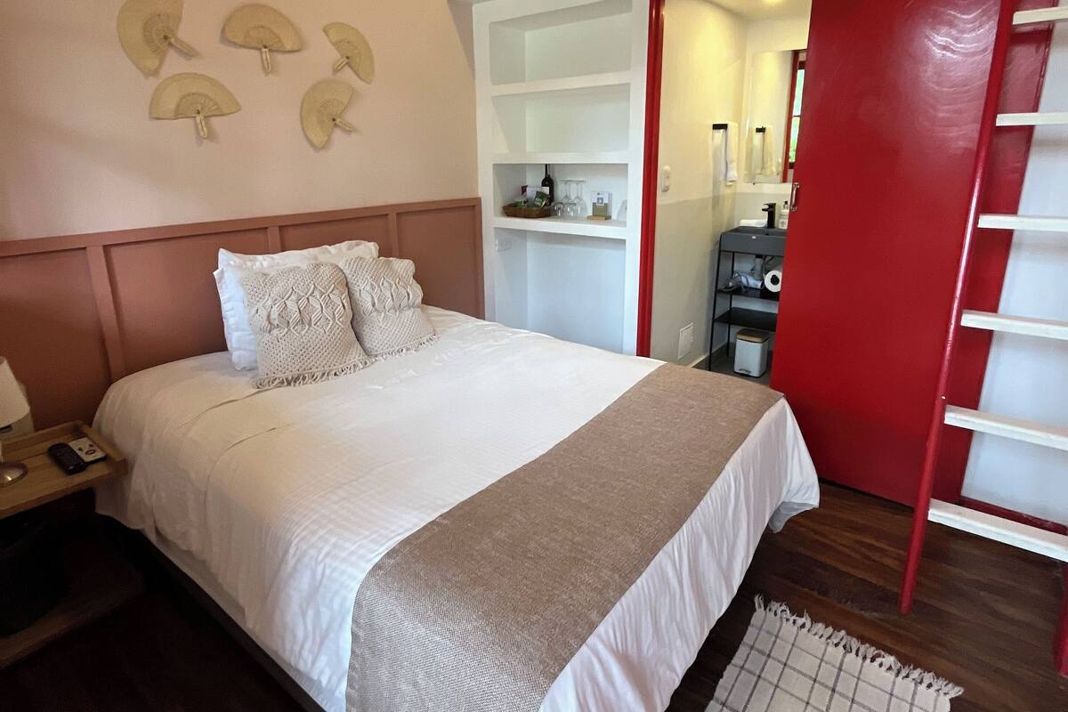 Quarto com cama, mesa de cabeceira e janela com moldura vermelha.