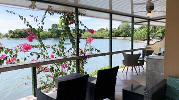 Terraço com mobiliário de vime com vista para um corpo d'água, teto de vidro e uma paisagem verde com um edifício ao longe.