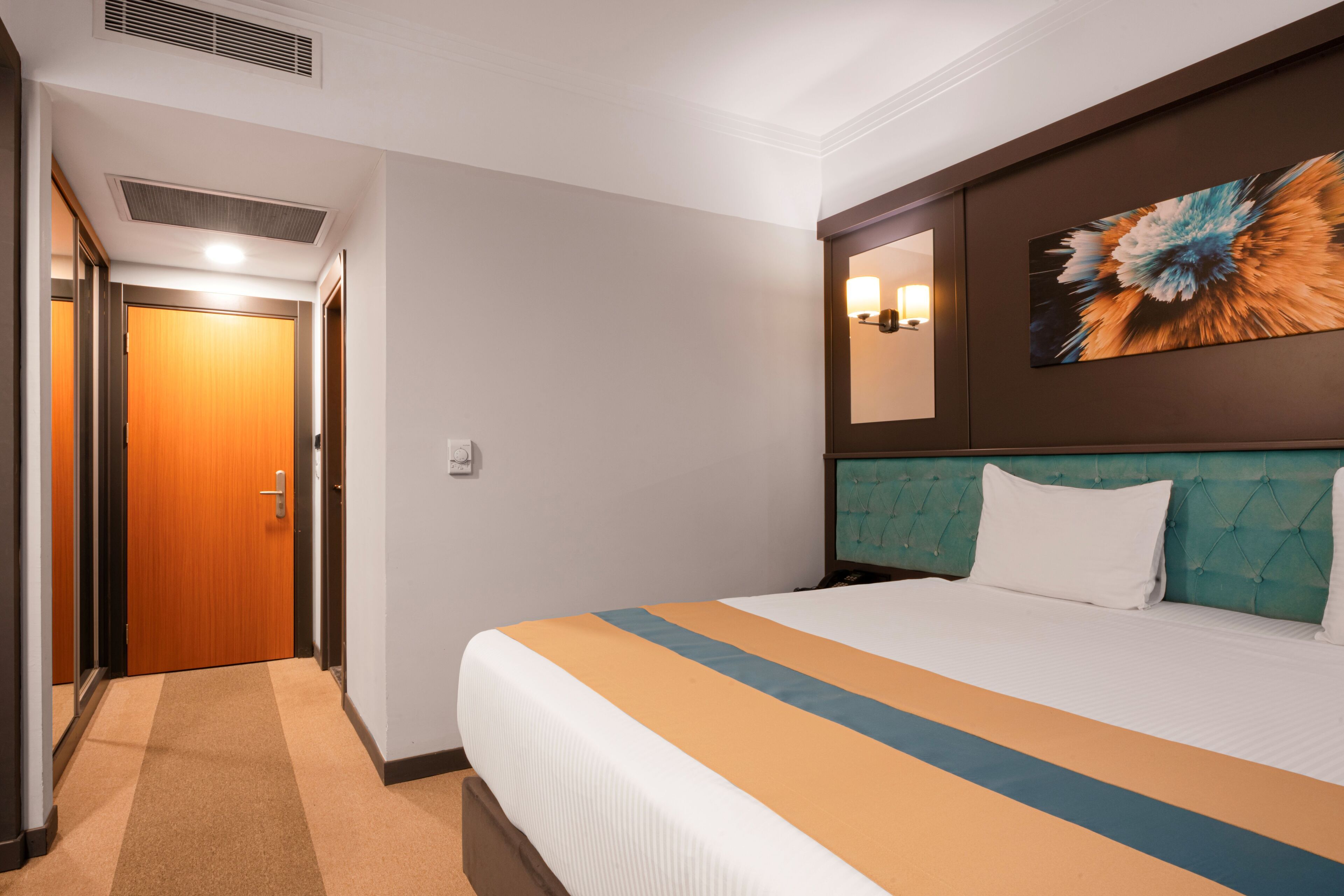 Quarto de hotel com uma cama, porta, luminária fixada na parede e um quadro.