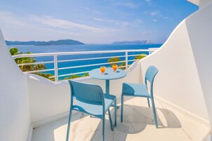 Varanda com duas cadeiras azuis e uma pequena mesa redonda, com vista para um mar azul e montanhas ao longe.