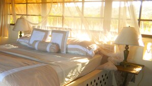 Uma cama com colcha e travesseiros brancos, uma mesa de cabeceira com abajur e uma janela com cortinas leves.
