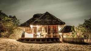 Uma cabana com telhado de palha, varanda e duas cadeiras, rodeada por areia e vegetação.