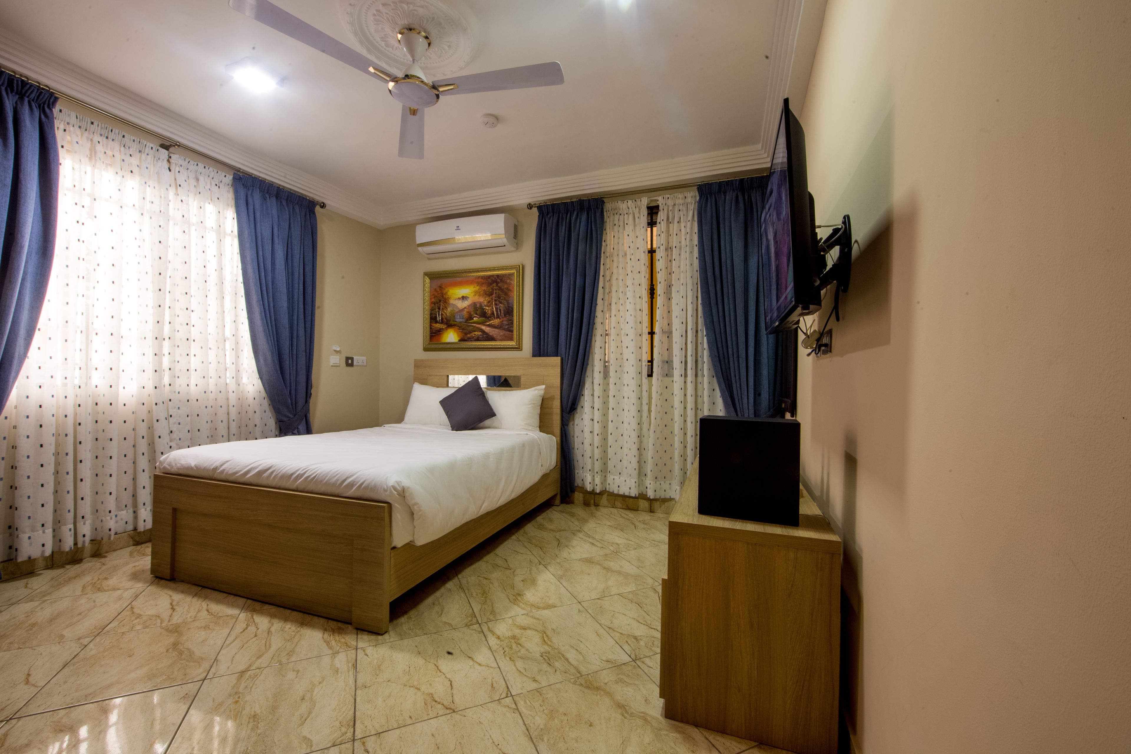 Quarto de hotel com cama, cortinas, ventilador de teto, ar condicionado, um quadro e uma TV sobre um suporte.