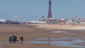 Uma praia com uma torre vermelha imponente ao fundo, um píer que se estende sobre a água e pessoas praticando diversas atividades.