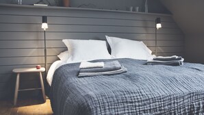 Cama bem arrumada com colcha cinza xadrez, duas almofadas brancas e um banquinho com um objeto vermelho ao lado.