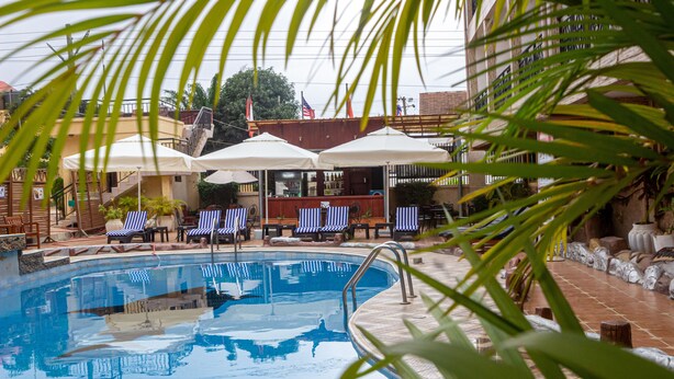 Uma piscina com espreguiçadeiras e guarda-sóis, rodeada por edifícios e palmeiras.
