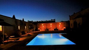 Piscina com espreguiçadeiras e área de estar, cercada por paredes com janelas e um muro.