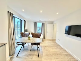 Uma sala de estar moderna com uma mesa de jantar, duas cadeiras, uma TV na parede e uma janela grande com cortinas.