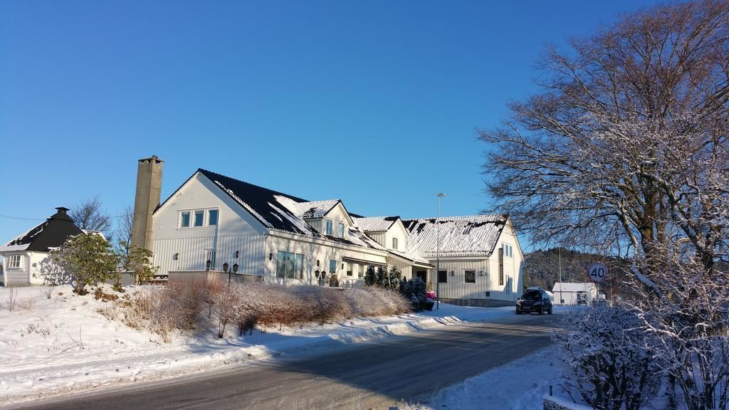 Uma casa grande e térrea, com telhado inclinado e chaminé, rodeada por neve no chão e árvores.