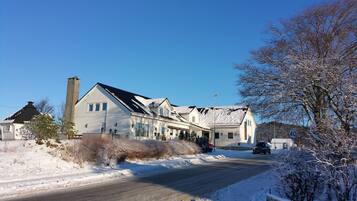 Uma casa grande e térrea, com telhado inclinado e chaminé, rodeada por neve no chão e árvores.