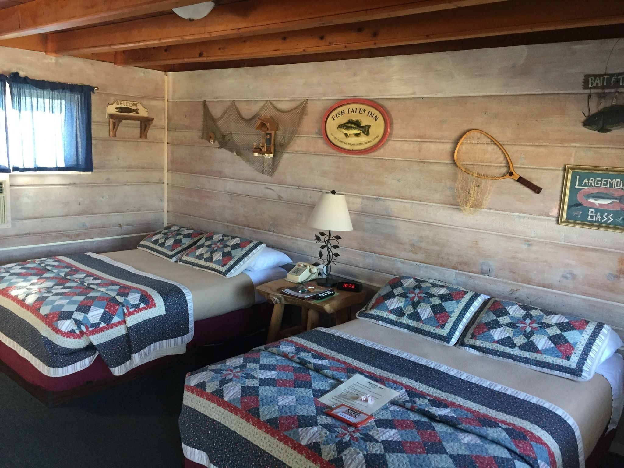 Quarto estilo cabana com duas camas, teto de madeira, placa de boas-vindas, placa com a inscrição 'histórias de peixes' e placa de anzóis e equipamentos de pesca.