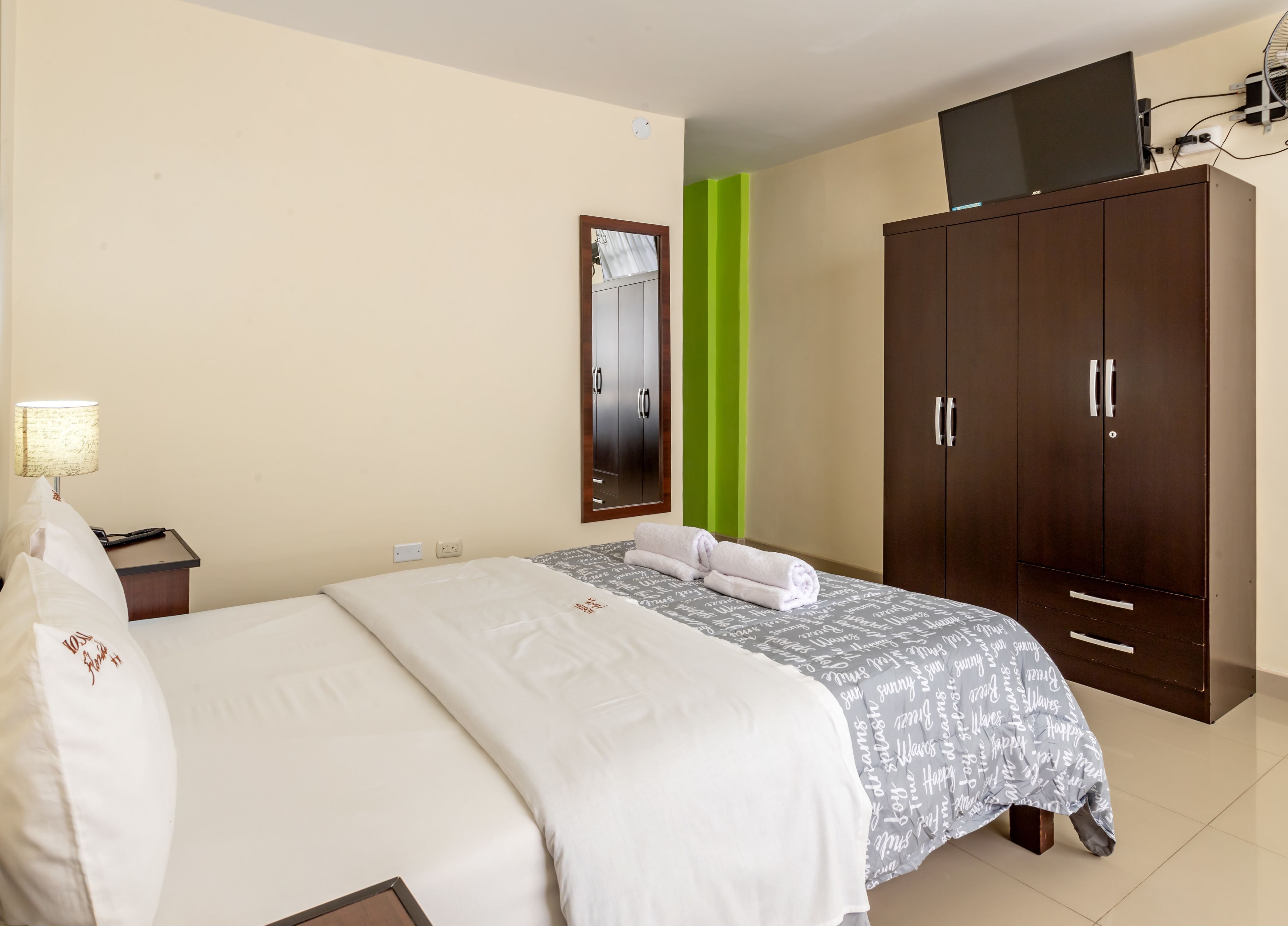 Quarto de hotel com cama, mesa de cabeceira, luminária, espelho, guarda-roupa e televisão.