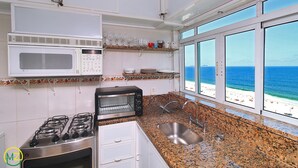Cozinha com fogão a gás, micro-ondas e forno elétrico, pia e janela com vista para o oceano.