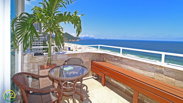 Uma varanda com móveis de vime, uma palmeira e vista para a praia e o oceano.