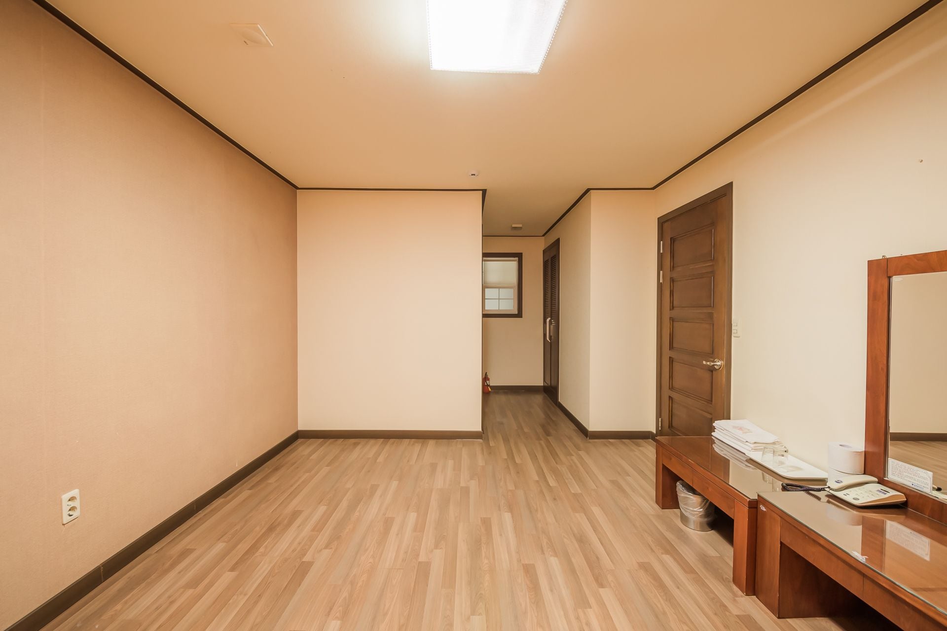 Um corredor com piso de madeira, um banco, uma mesa com telefone e uma porta.