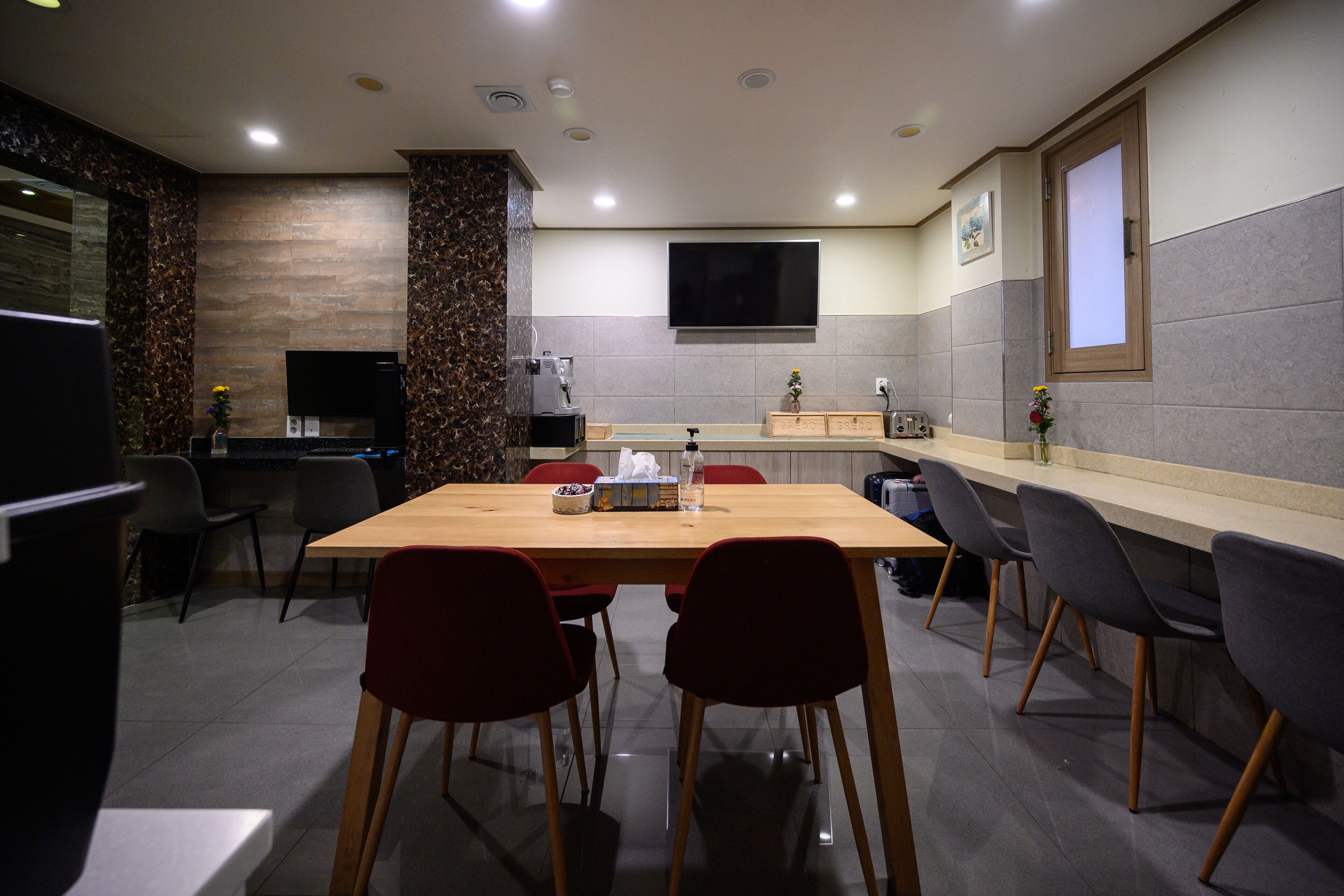 Uma zona de refeições moderna com mesa de madeira, cadeiras vermelhas e cinzentas, área de cozinha com televisão e uma janela ampla.
