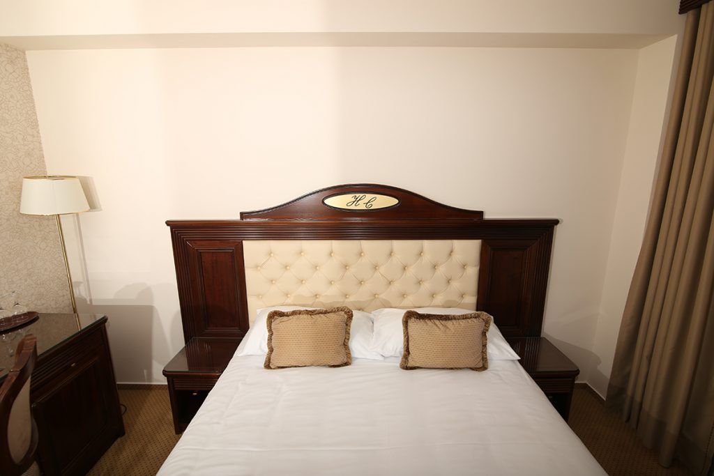 Quarto de hotel com uma cama grande, cabeceira de madeira, duas mesas de cabeceira, uma luminária e uma janela com cortinas.
