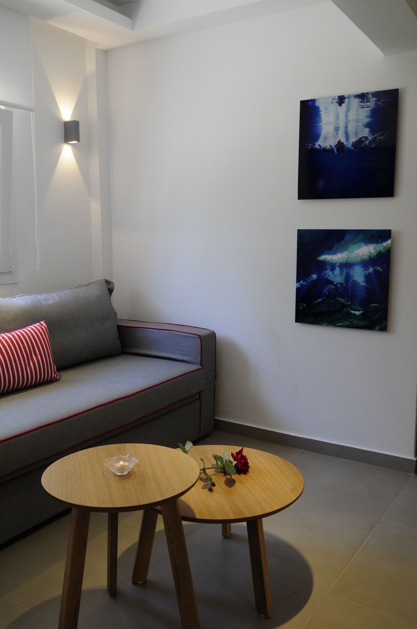 Uma sala de estar moderna com um sofá cinzento, uma mesinha de madeira com uma vela e flores, e duas pinturas abstratas na parede.