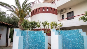 Área de piscina com azulejos azuis em mosaico, uma palmeira e um edifício branco com detalhes vermelhos.