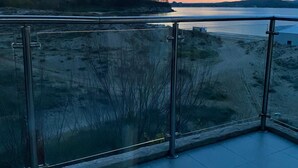 Varanda com guarda-corpo de vidro, vista para uma praia e para o mar ao pôr do sol.