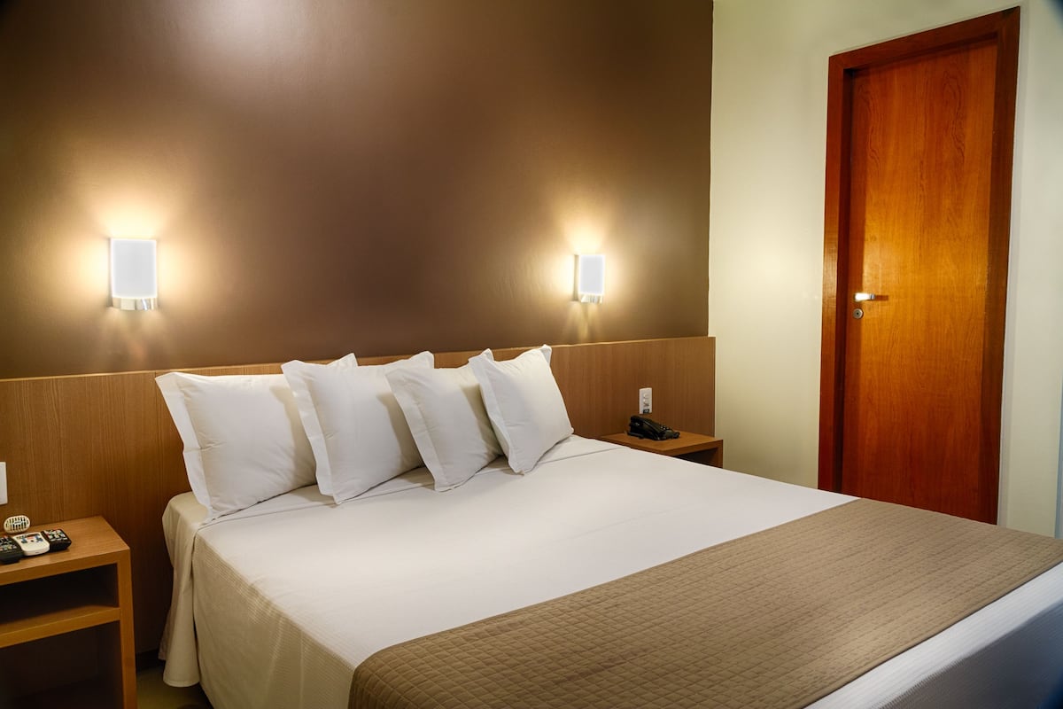 Quarto de hotel com uma cama grande, duas mesas de cabeceira, um painel de cabeceira de madeira e duas luminárias fixadas na parede.