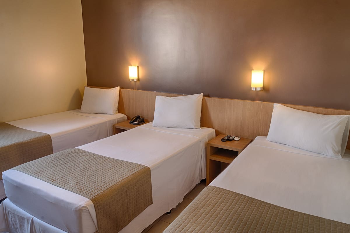 Quarto de hotel com três camas de solteiro, cada uma com mesa de cabeceira e luminária.