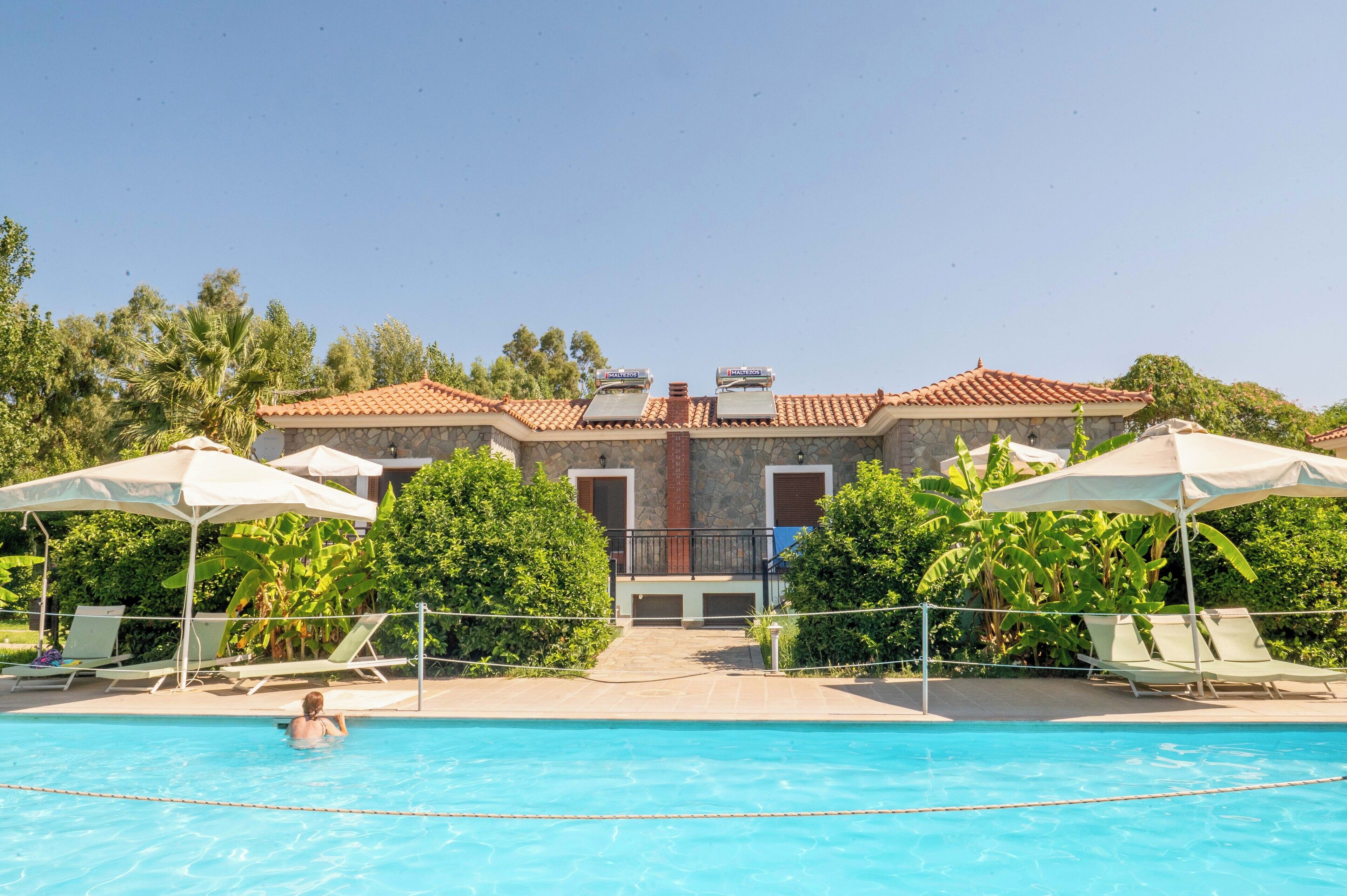 Piscina com espreguiçadeiras e guarda-sóis, cercada por vegetação e um edifício com telhado em azulejos.