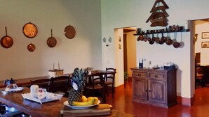 Uma área de refeições com uma mesa de madeira posta com uma abacaxi e bananas, uma cafeteira e um tabuleiro branco. Cadeiras cercam a mesa, e um armário de madeira com um espelho e prateleiras acima.